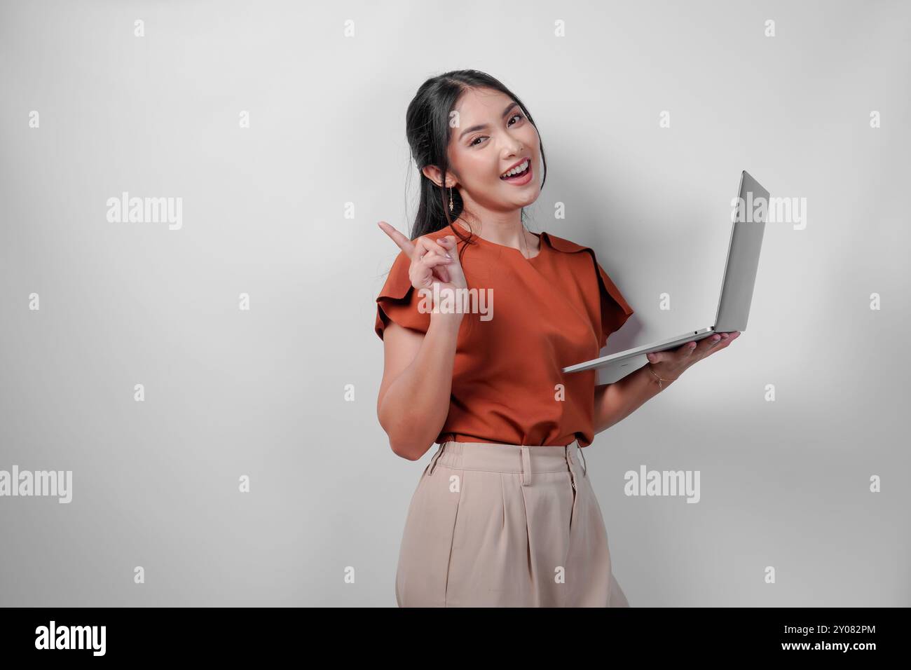 Confident young Asian woman in brown shirt holding laptop while pointing to the copy space ...
