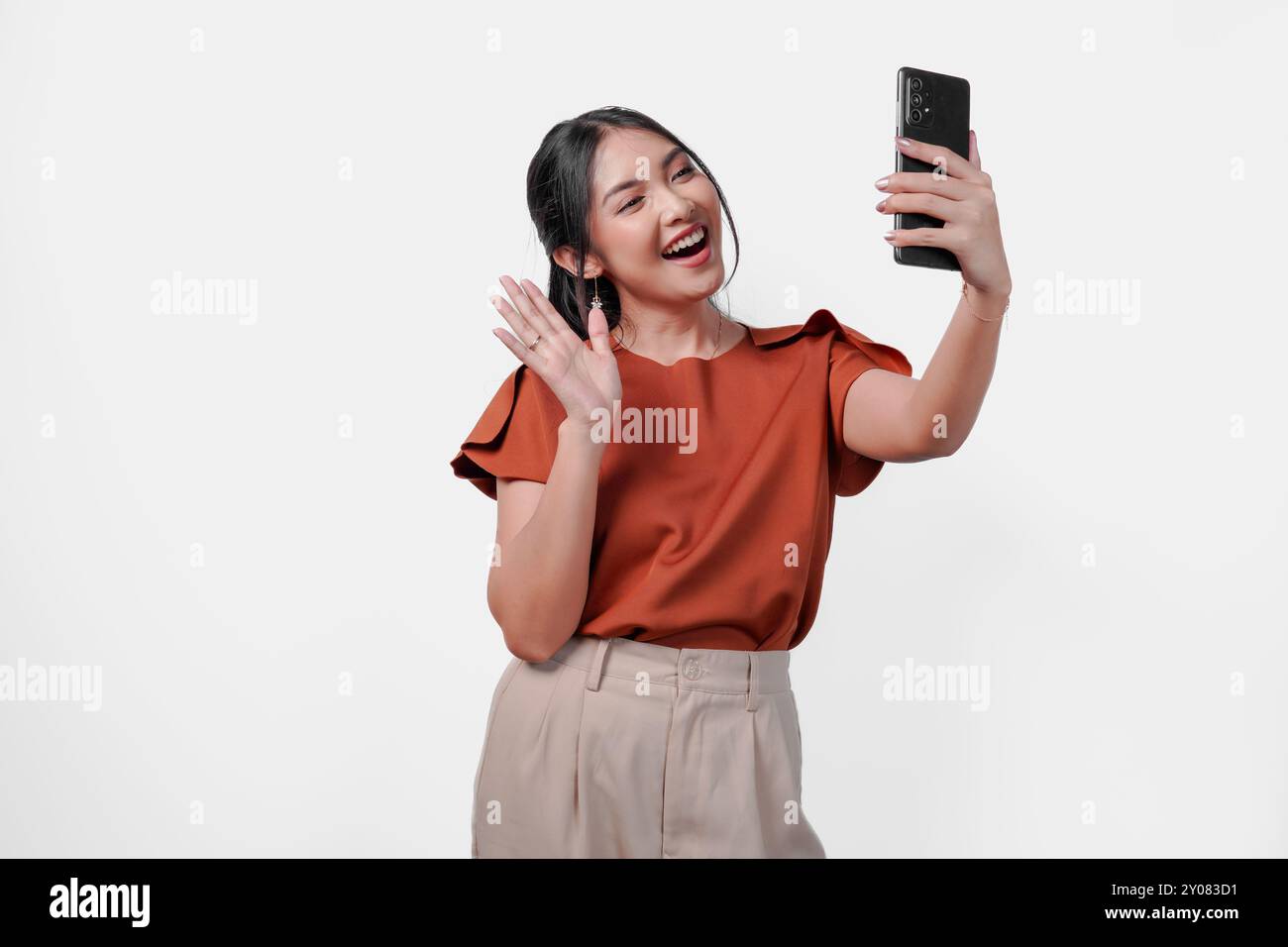 Smiling Asian woman in brown t-shirt doing video call pose while waving ...