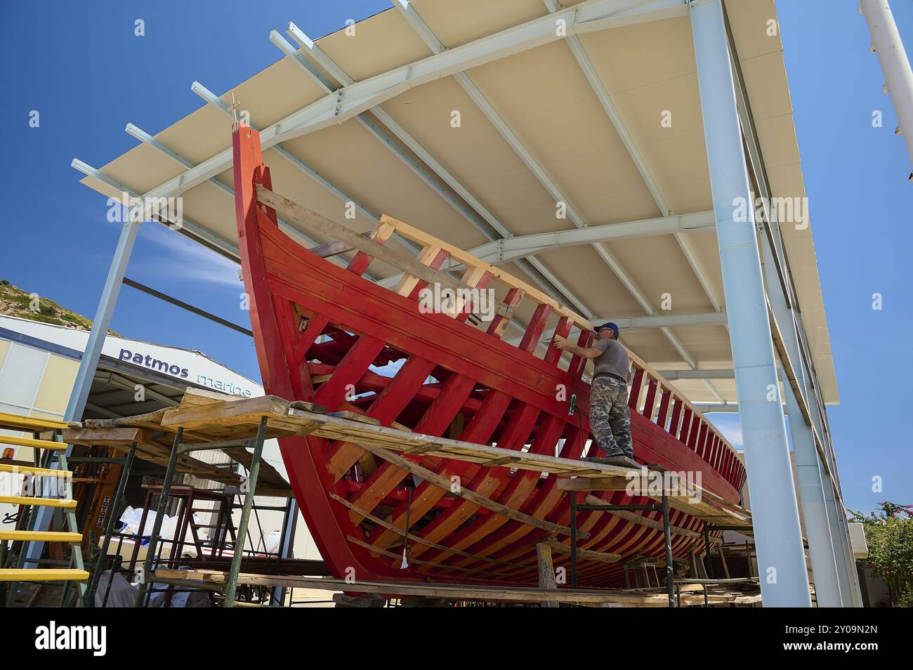 A partially built red wooden ship in a covered shipyard with a worker ...
