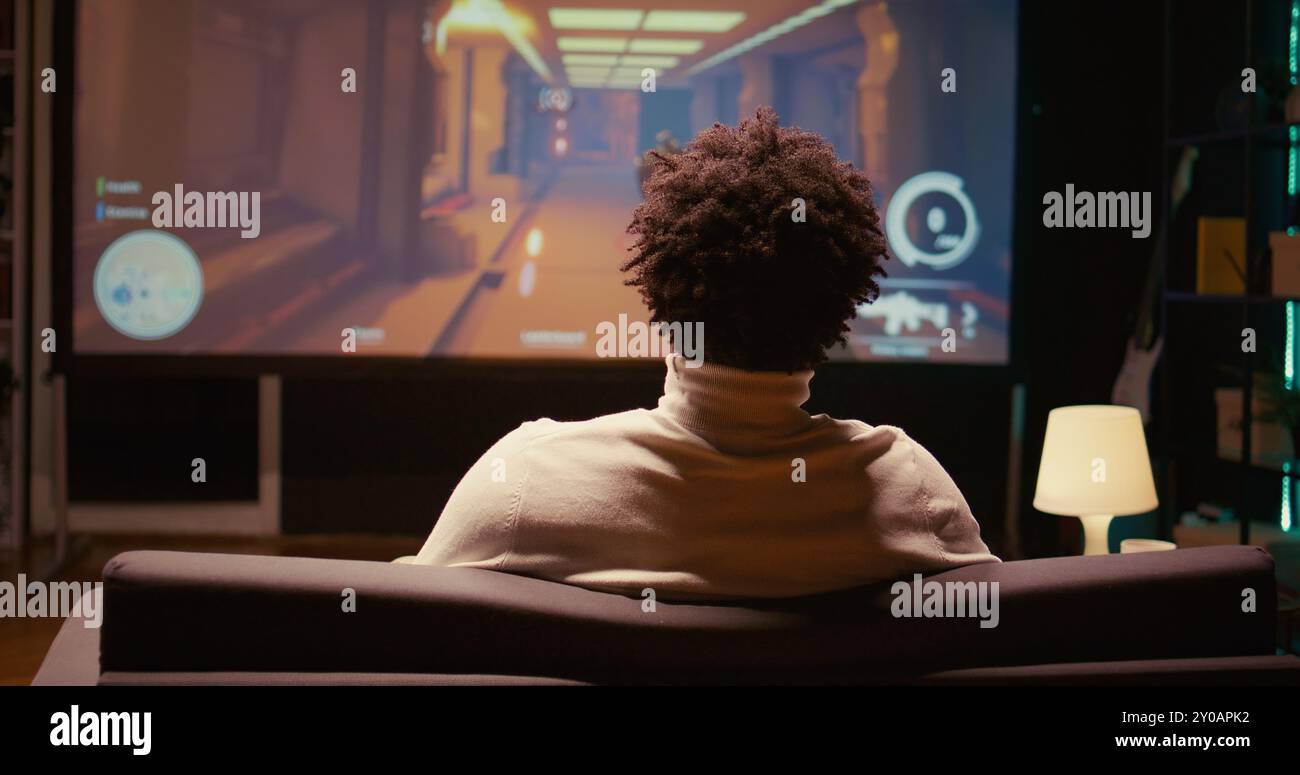 BIPOC man playing shooter videogame on ultrawide smart TV in futuristic industrial setting with ...