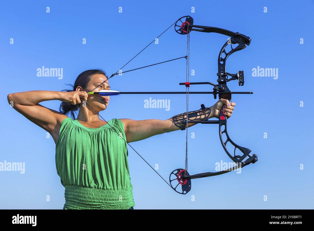 Young black haired colombian woman aiming arrow of compound bow in blue ...