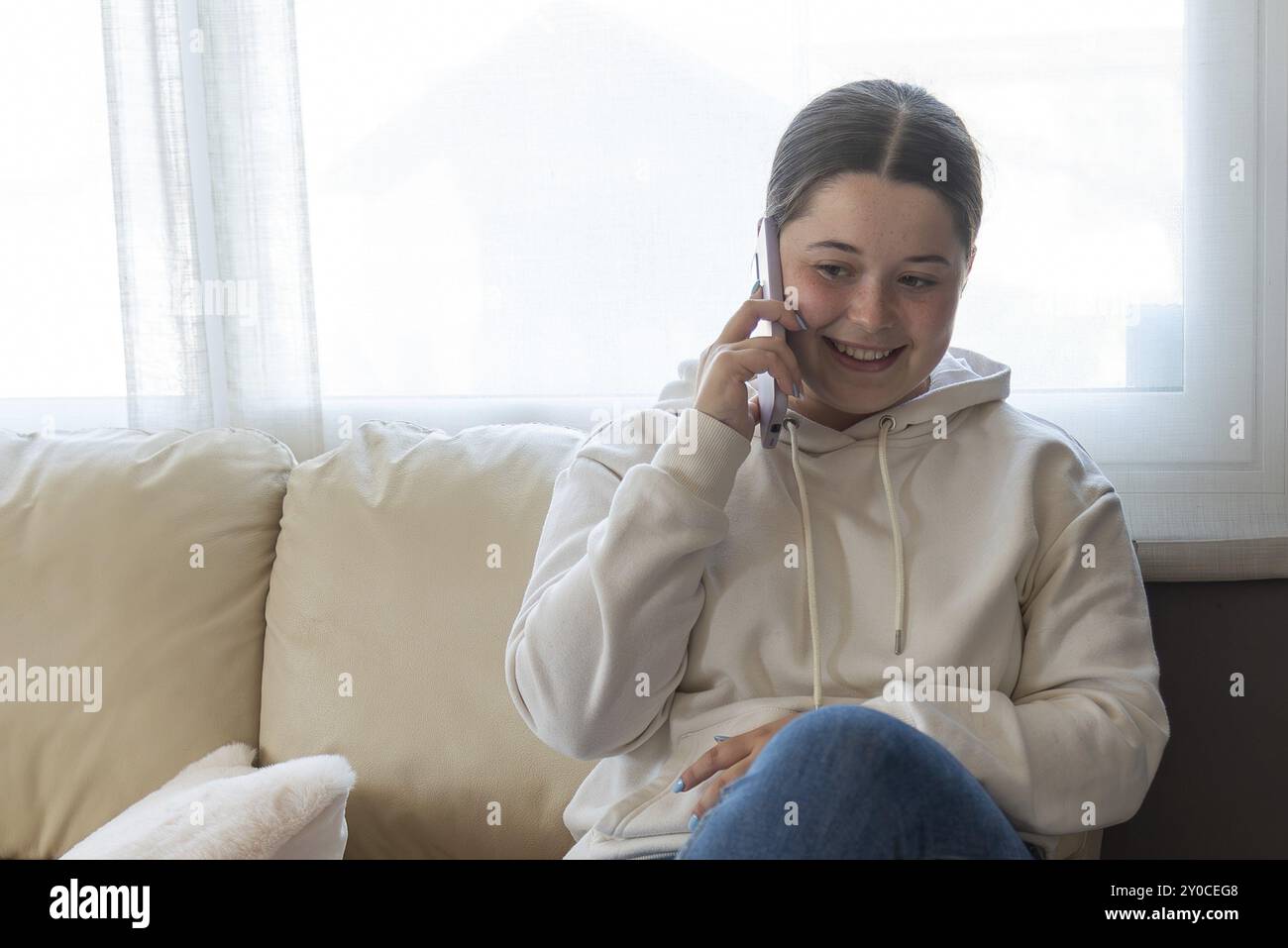 Young woman sitting on a beige sofa, enjoying a phone conversation in a bright, homey space ...