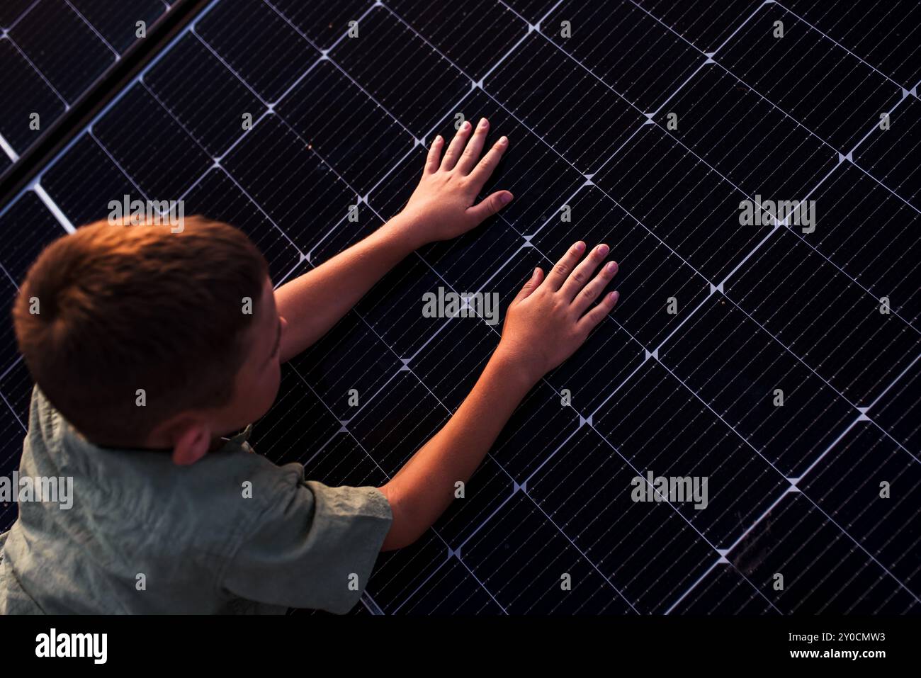 Boy lying on belly on roof with solar panels, touching panel with hands ...