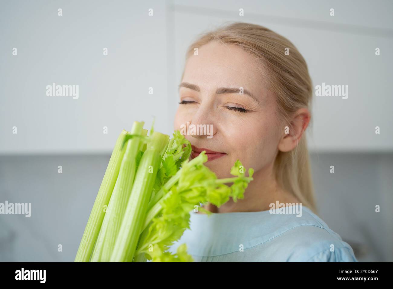 A woman is savoring fresh celery in a modern kitchen, embodying a healthy lifestyle concept ...