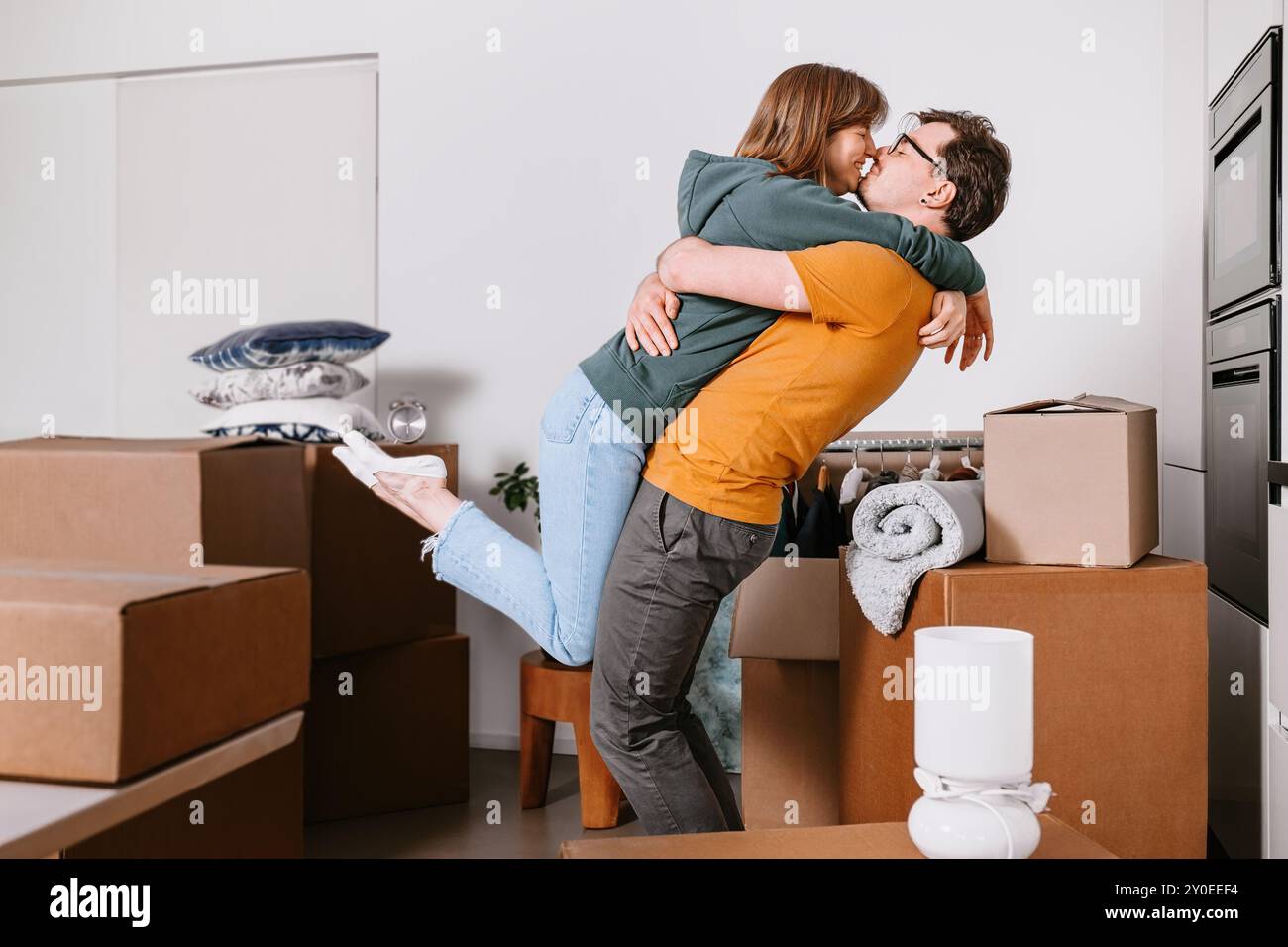 Couple is playfully hugging and lifting each other in new home, surrounded by moving boxes ...