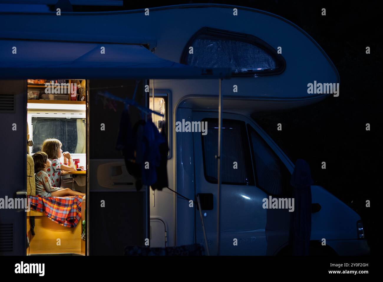Two kids, not looking, sitting in a camper van at night, seen from ...