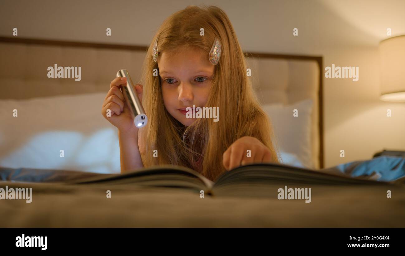 Curious Caucasian child little girl reading book instructive story textbook daughter lying bed ...