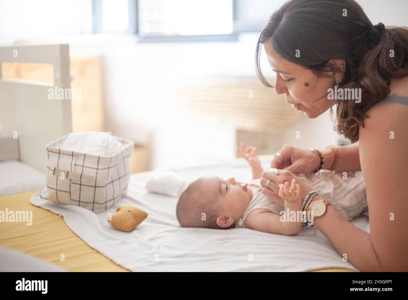 A woman fastens the buttons of her baby's dress on a bed at nappy ...