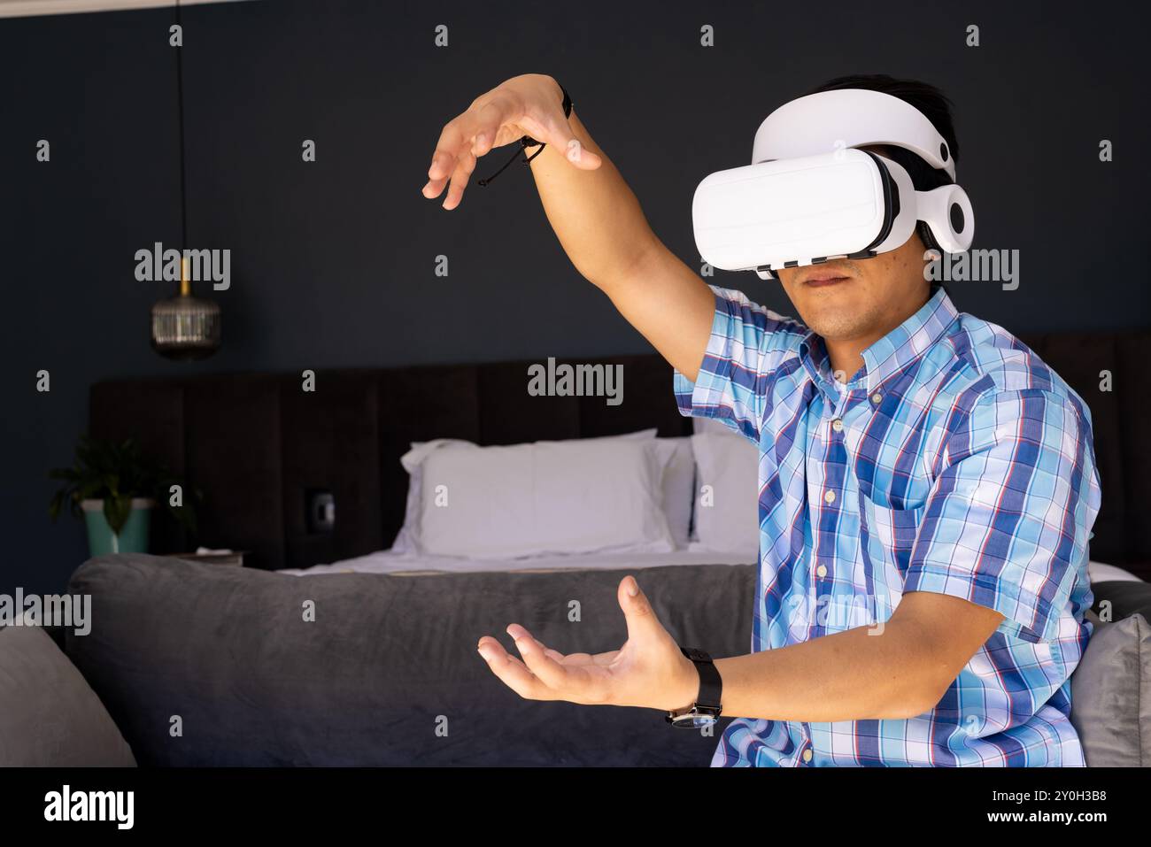Using VR headset, asian man in plaid shirt experiencing virtual reality in bedroom, copy space ...