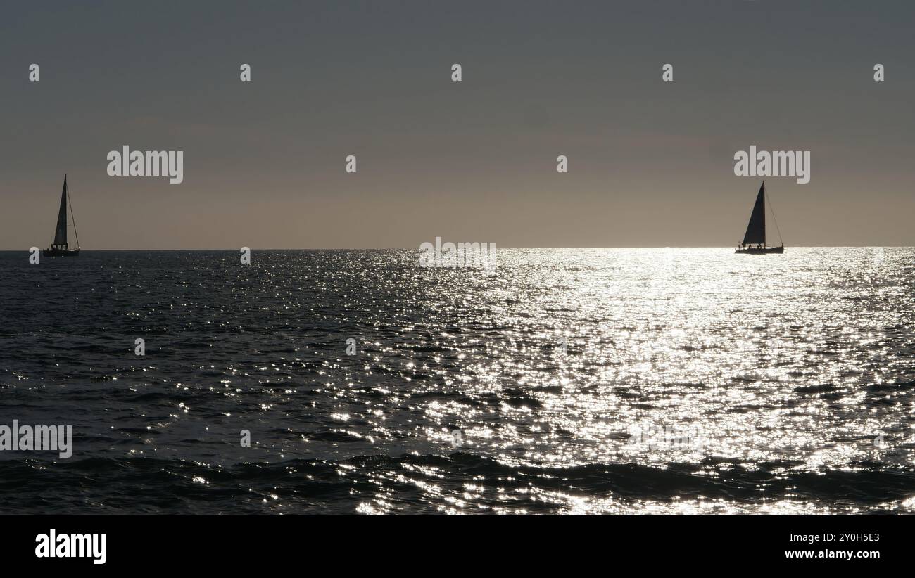 Two sailboats sailing away on calm ocean at sunset with sparkling water ...
