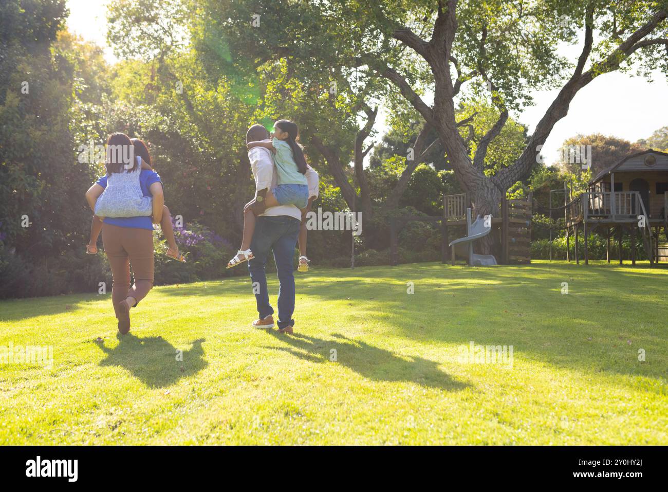 family carrying daughters on backs, walking together in sunny backyard ...