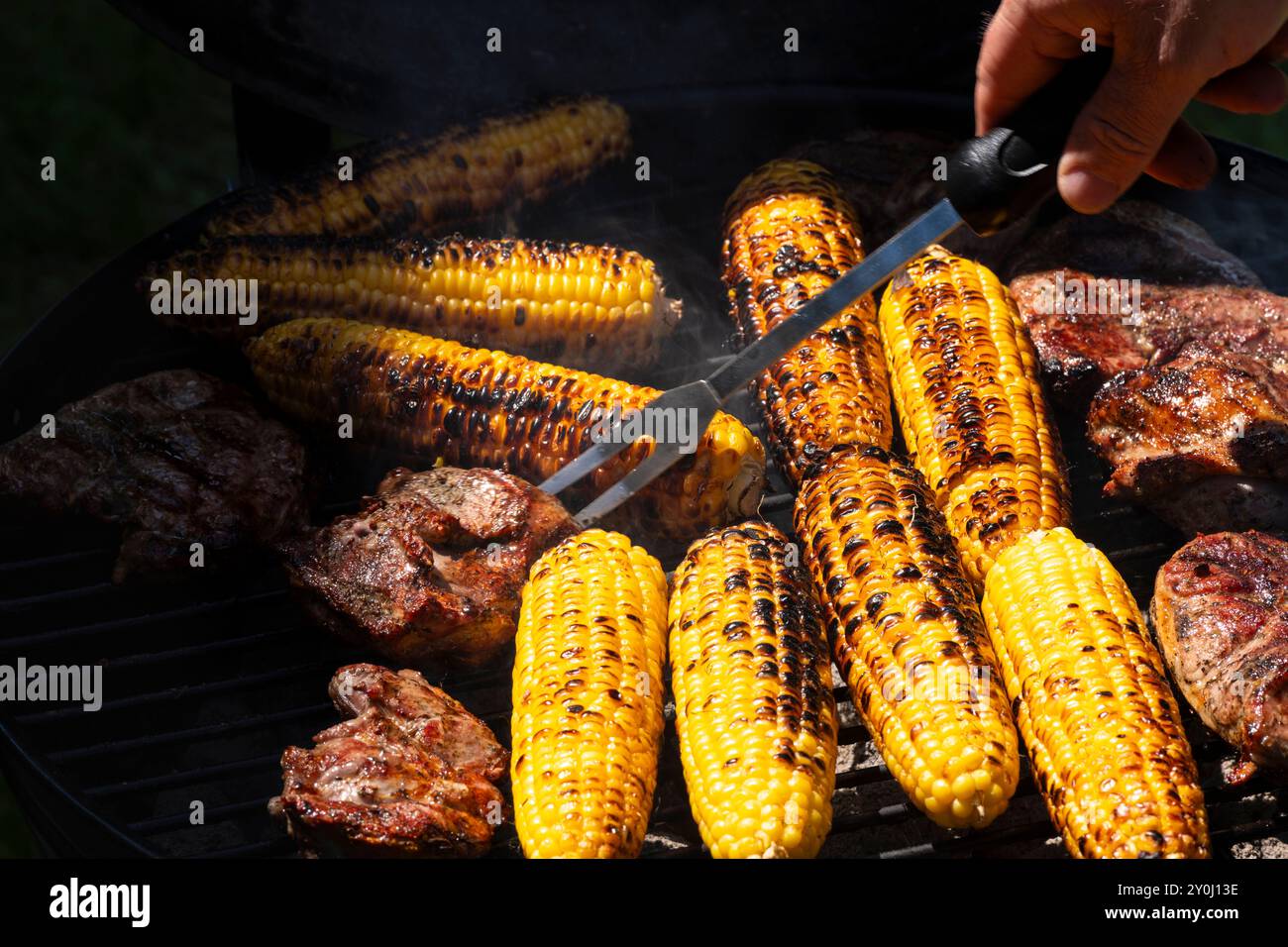 A hand flips meat on a barbecue grill, with golden, charred corn beside ...