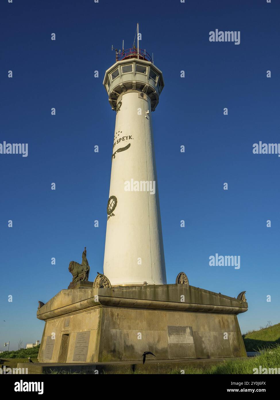 A tall lighthouse stands under a clear blue sky, with a monument below, egmond aan zee ...