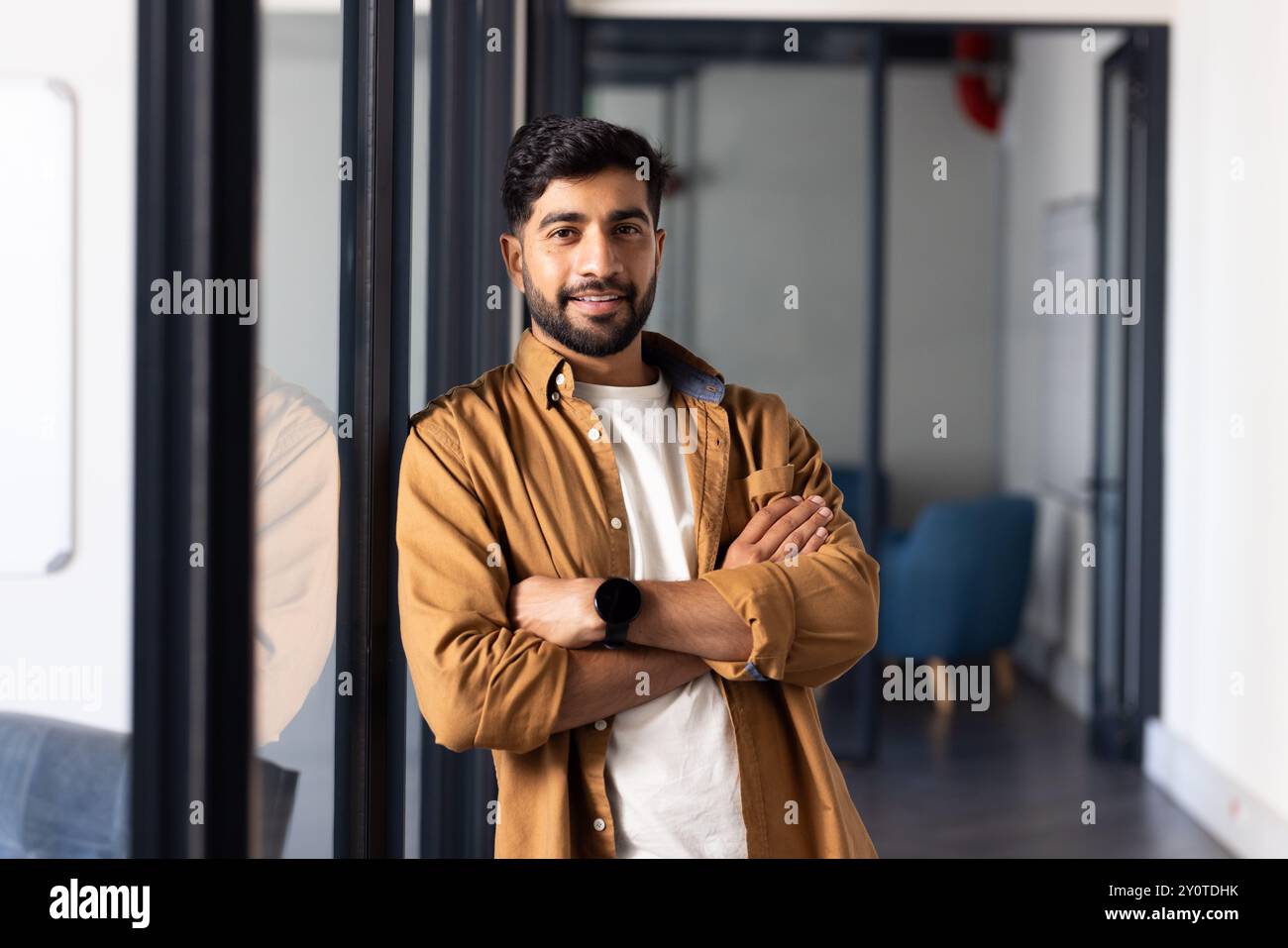 Smiling Indian man with crossed arms standing in modern office hallway ...