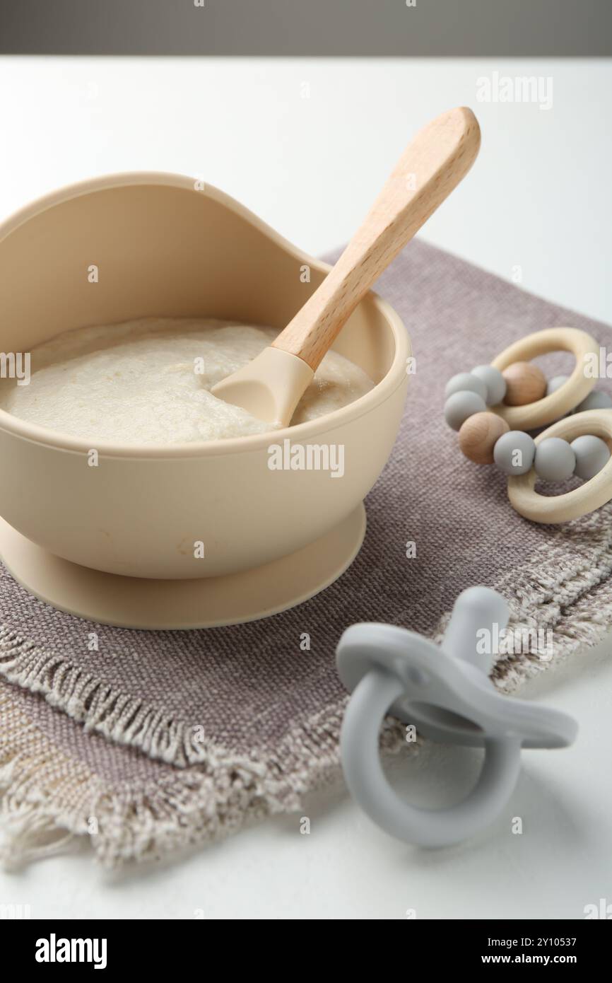 Delicious baby food in bowl with spoon, pacifier and teether on white ...