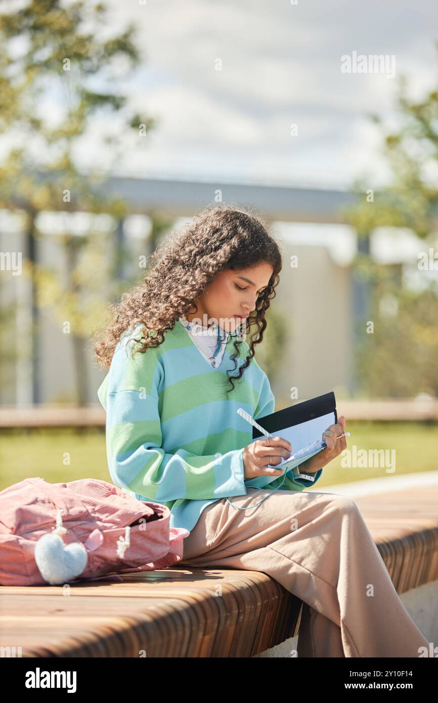 Curly teenage girl on bench writing in notebook doing homework while ...