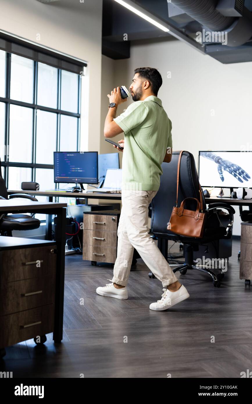 Walking in office, Indian man drinking coffee and holding tablet near ...