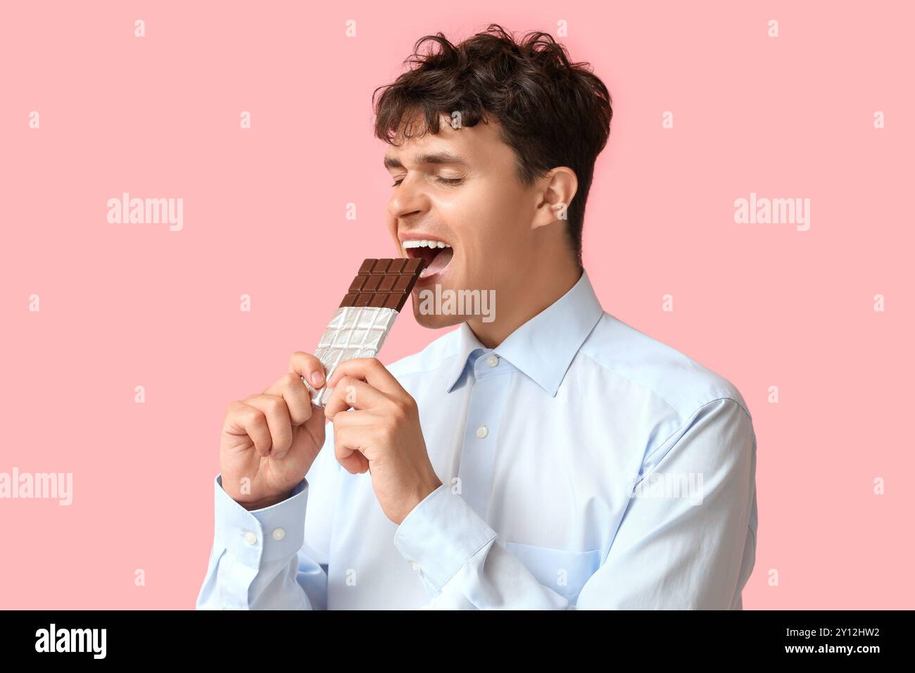 Handsome young man eating sweet chocolate bar on pink background Stock ...
