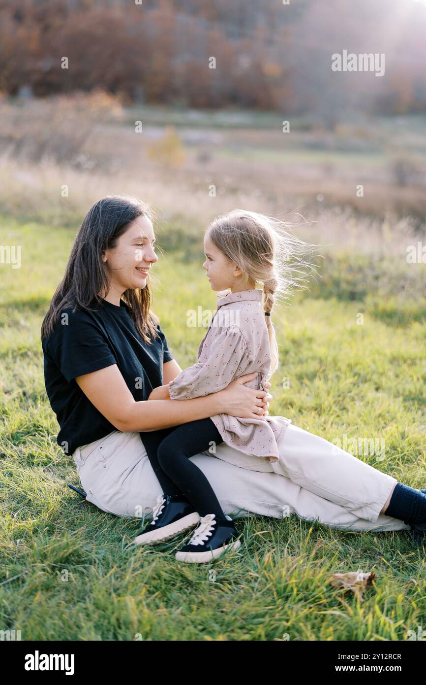 Mom looks at little girl hugging her sitting on her lap on green lawn ...