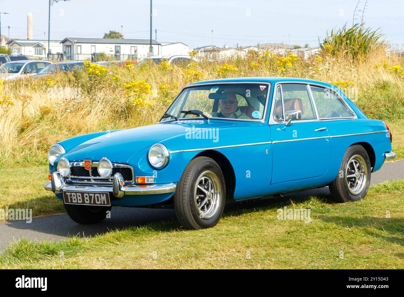 A vintage blue MGB GT car is parked on a grassy area, with a backdrop ...