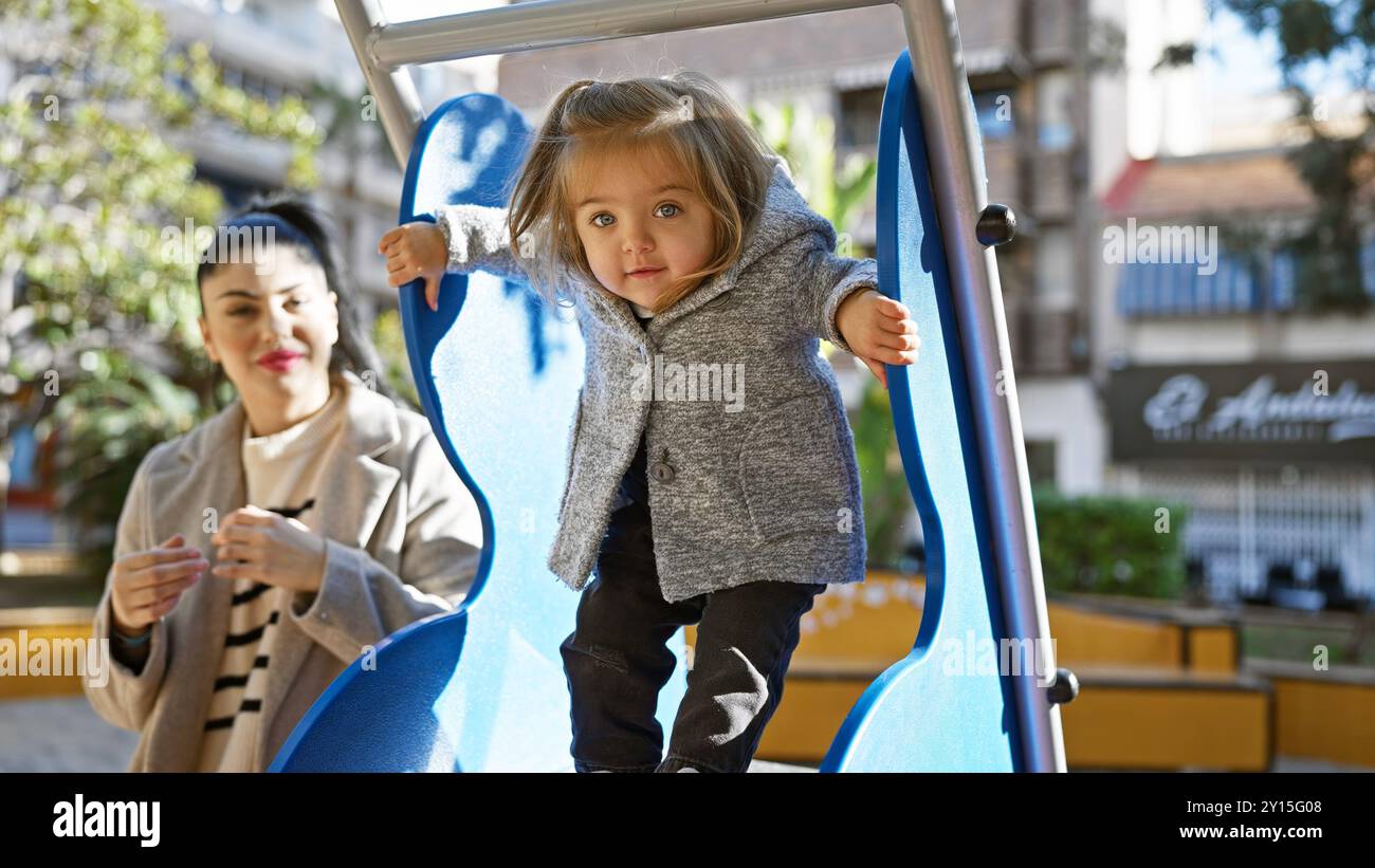 A young girl slides at a playground with her mother watching, capturing ...