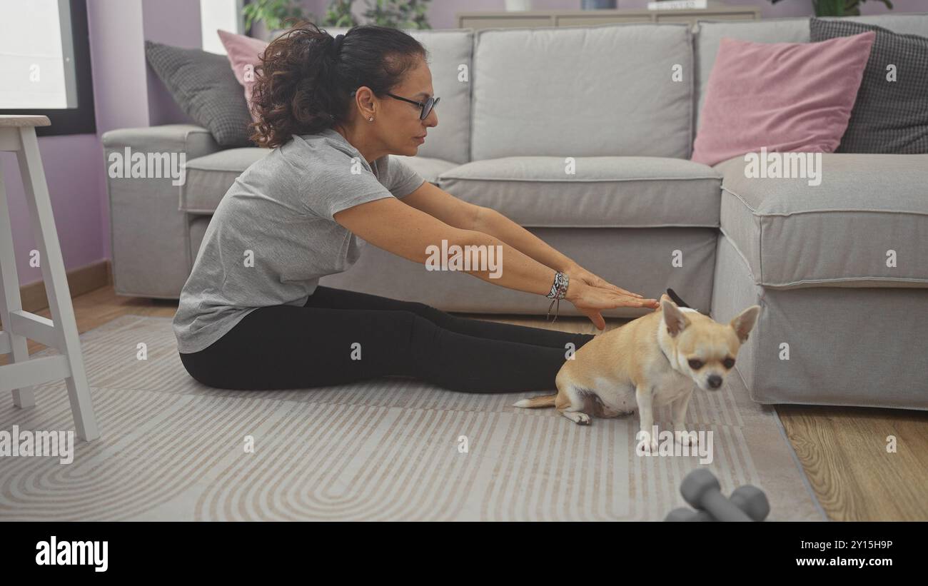 A middle-aged hispanic woman stretches at home with her chihuahua dog in a cozy living room ...