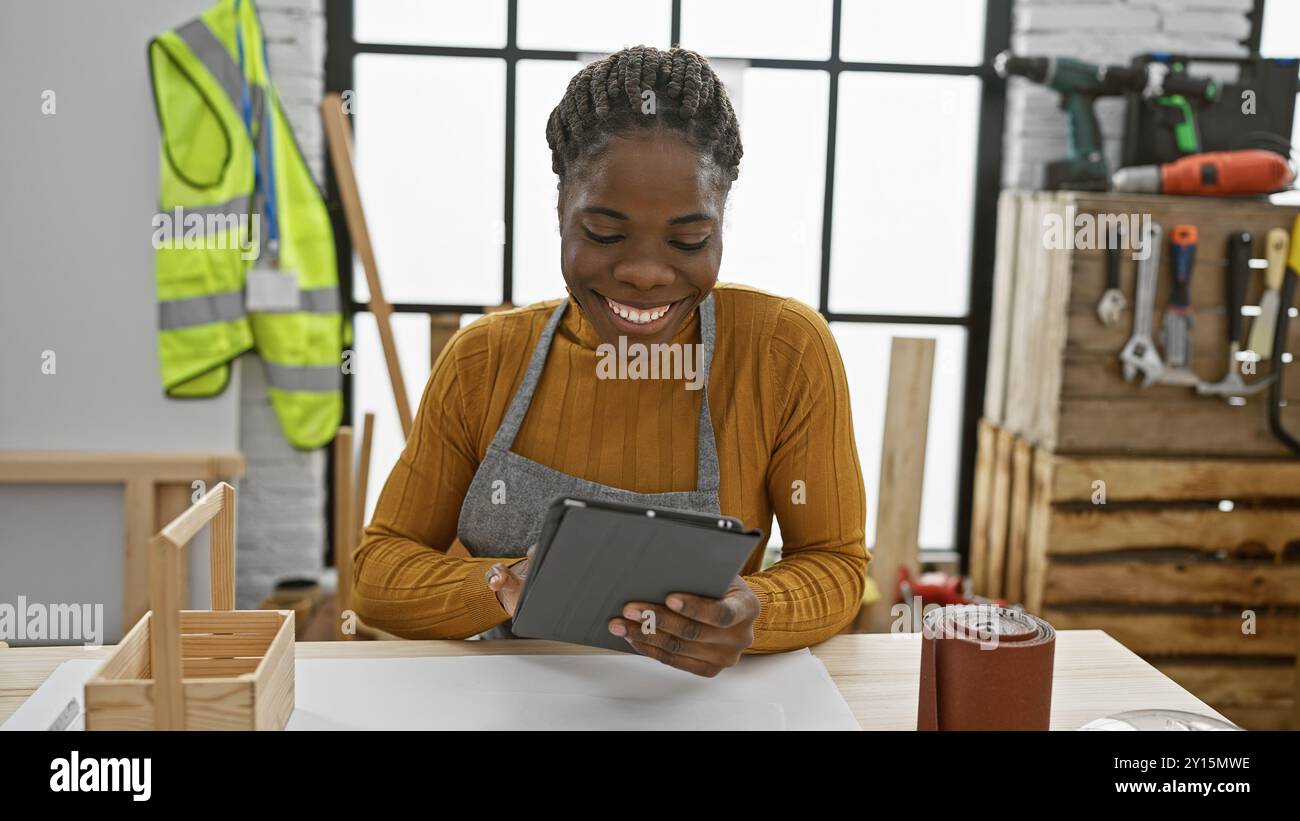 Smiling black woman with braids using tablet in carpentry workshop ...