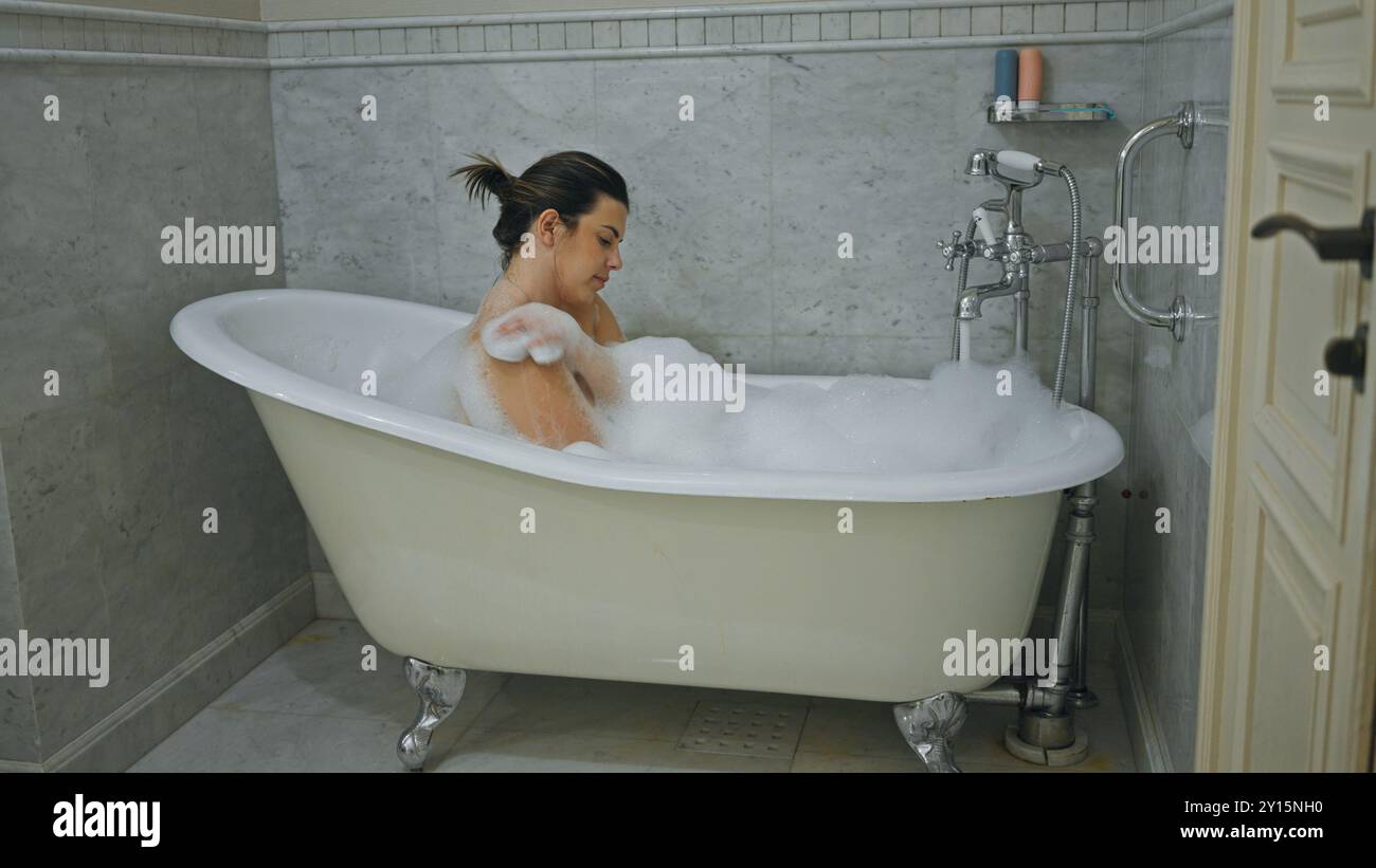 A relaxed young woman enjoying a bubble bath in a vintage-style bathtub ...