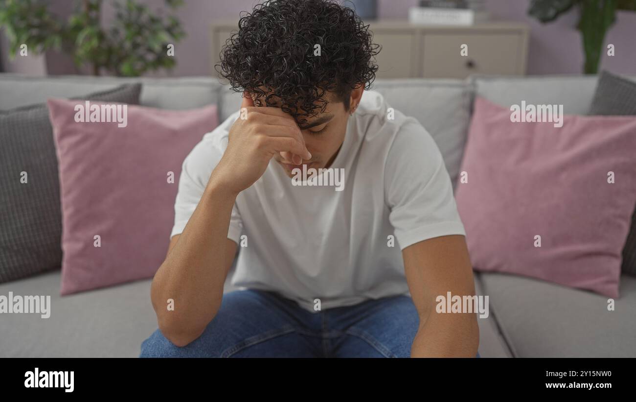 A distressed young hispanic man sitting on a grey couch indoors with ...