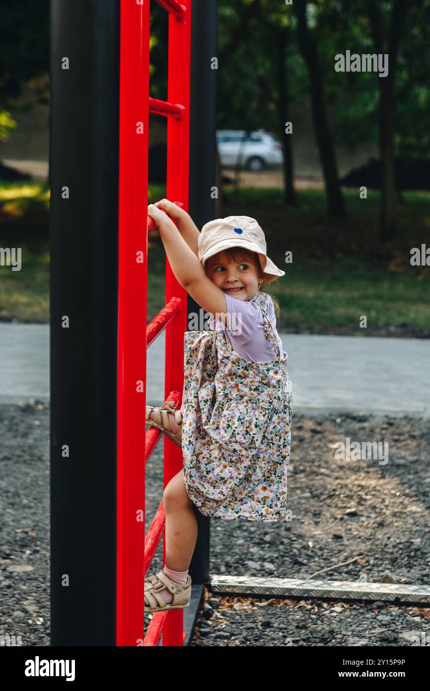 Young girl in a floral dress and sunhat climbing a red ladder structure ...