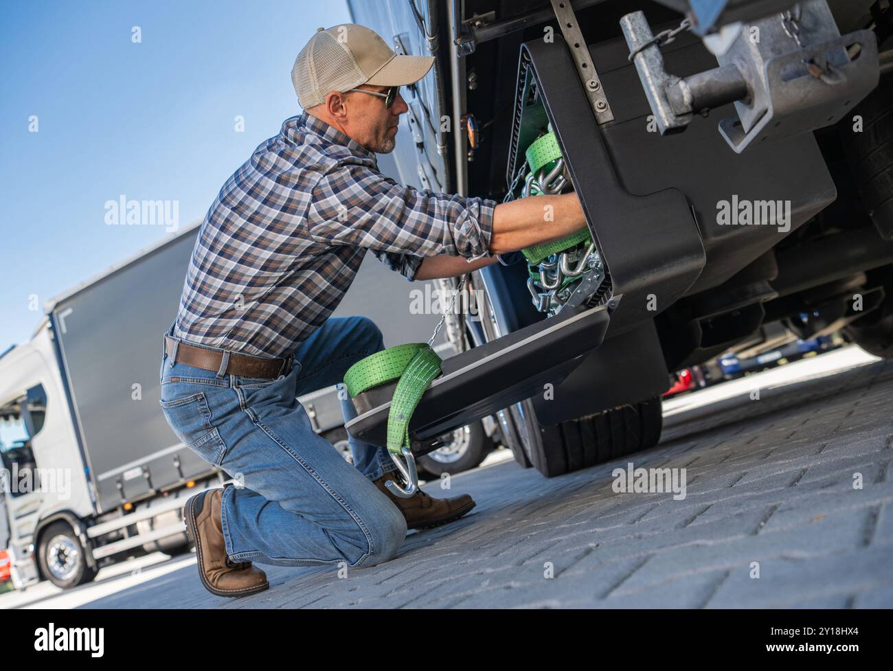 A truck driver is kneeling to secure cargo with green straps at a ...