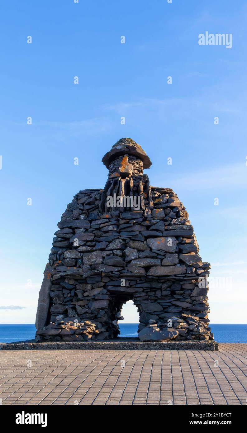 A large stone statue of a man stands in front of a blue sky. The statue ...