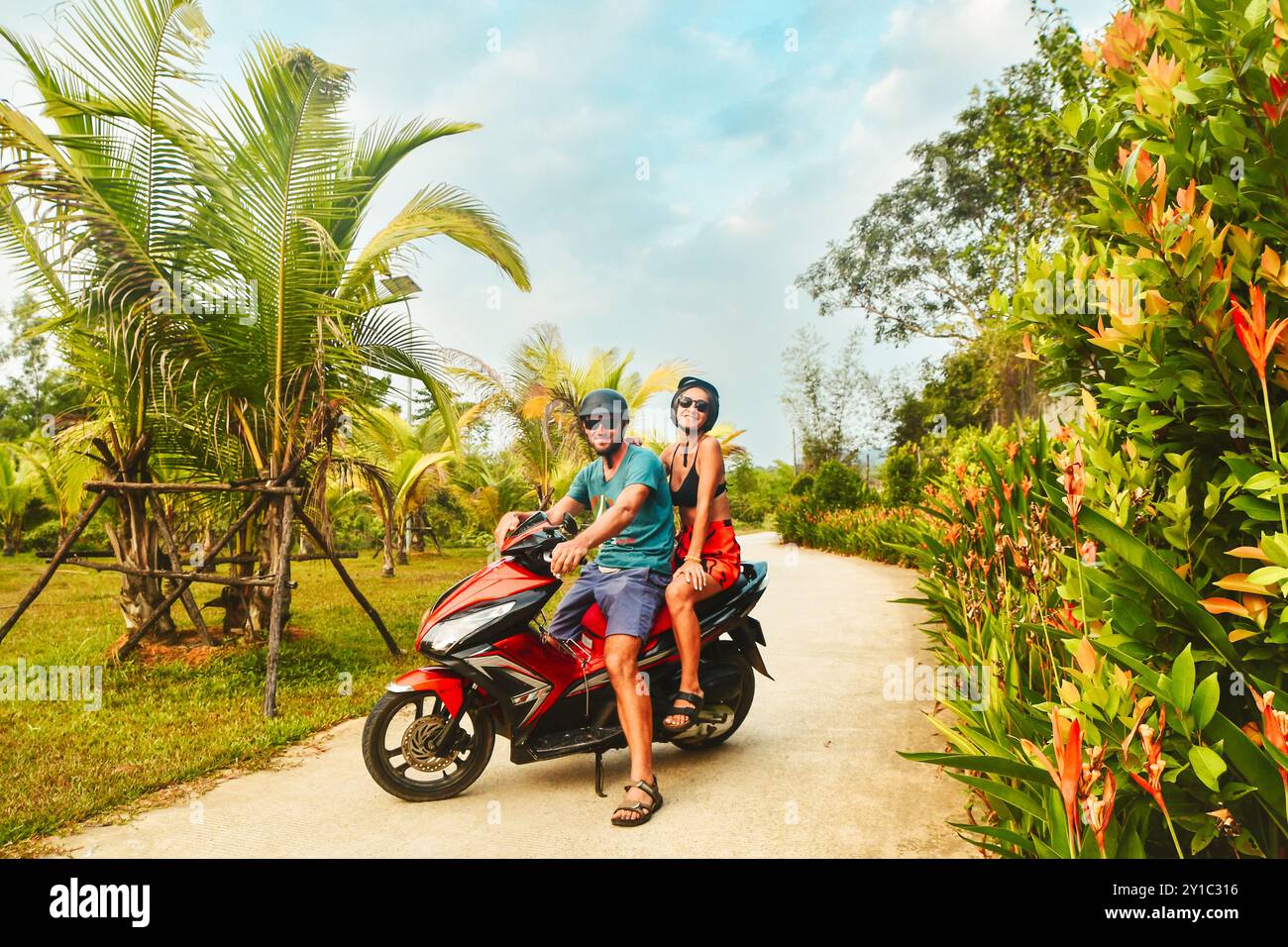 Happy smiling couple travelers riding motorbike scooter in safety helmets during tropical ...