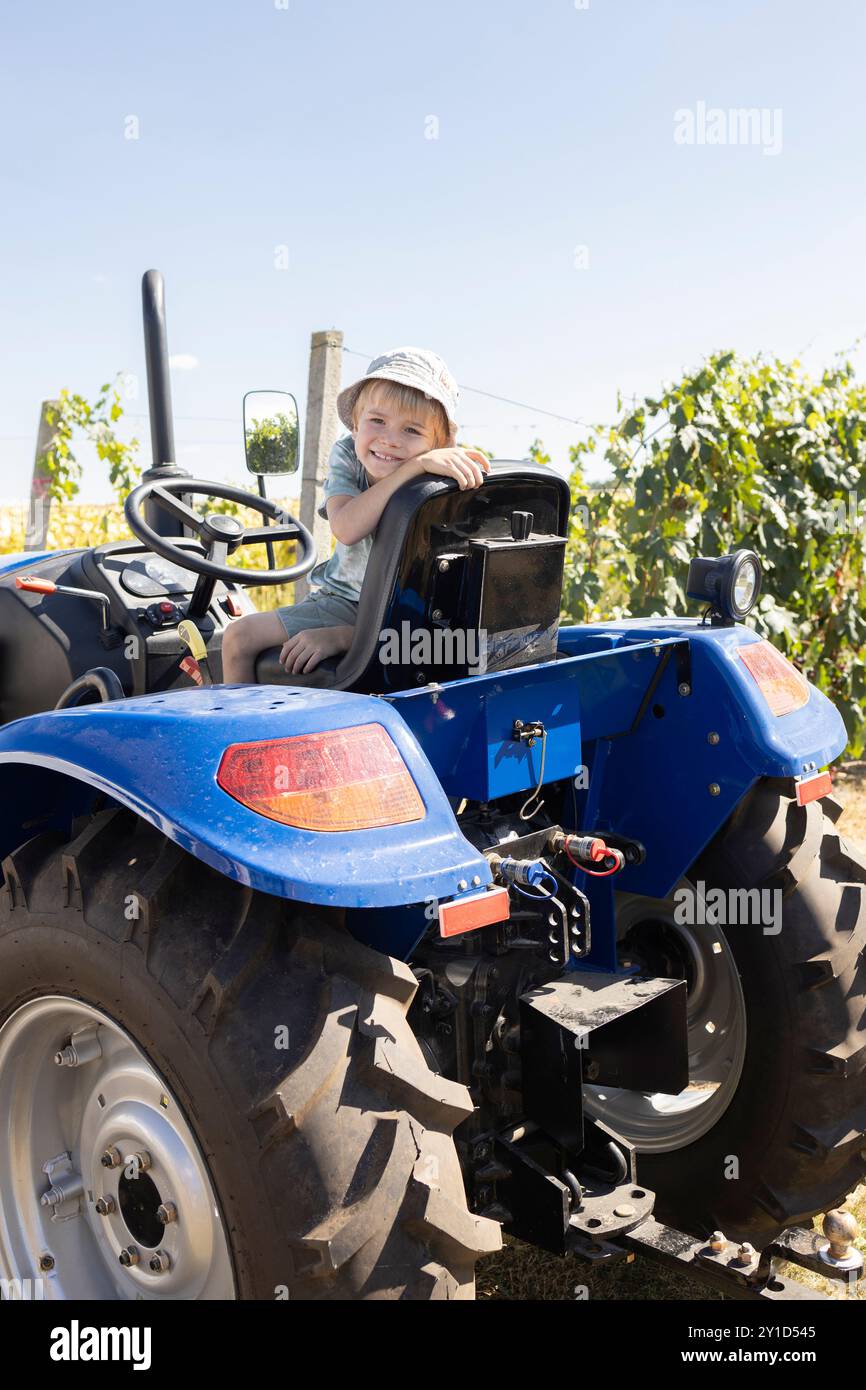 little blond boy happily sits behind the wheel of a tractor with big ...