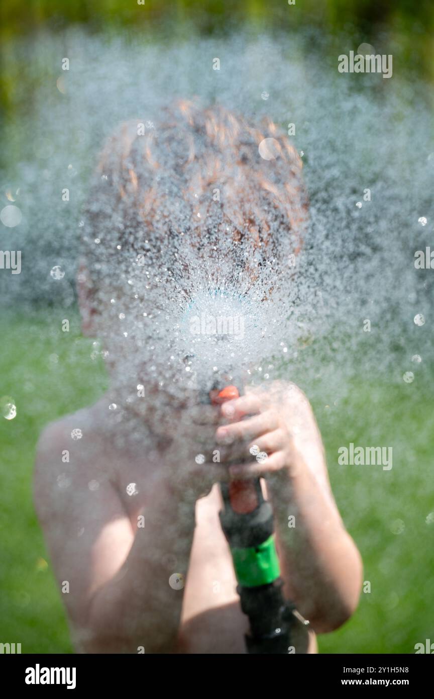 Little boy spraying water from a hose at a camera. In backyard during a ...