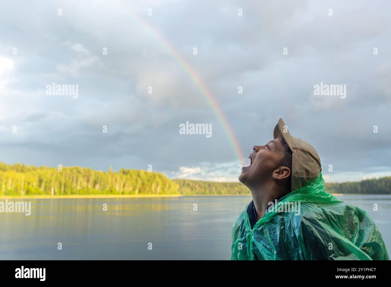 A boy playfully interacts with a rainbow against a serene lakeside ...