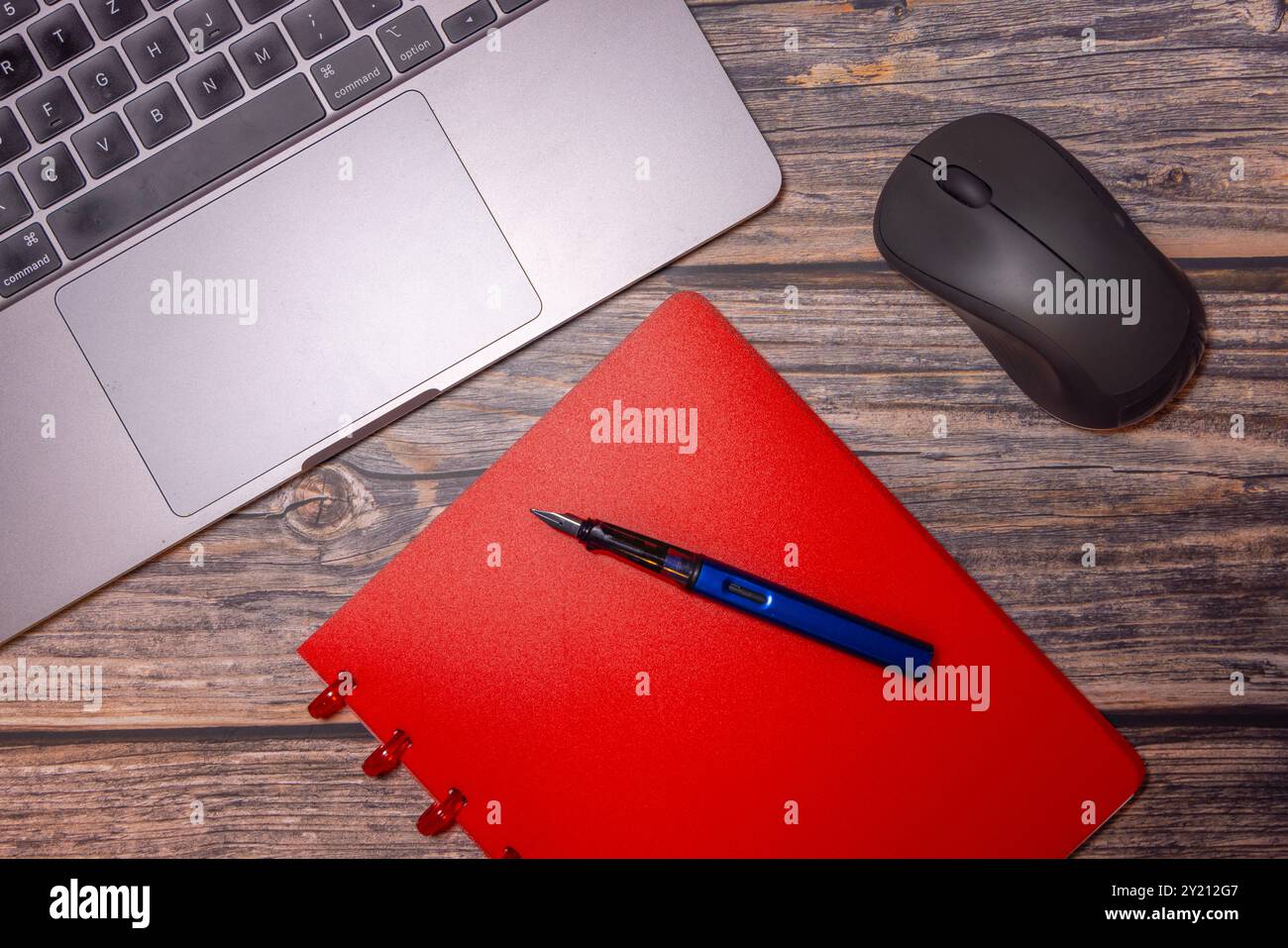 A flat lay composition featuring a red notebook, fountain pen, laptop, and mouse on a rustic ...