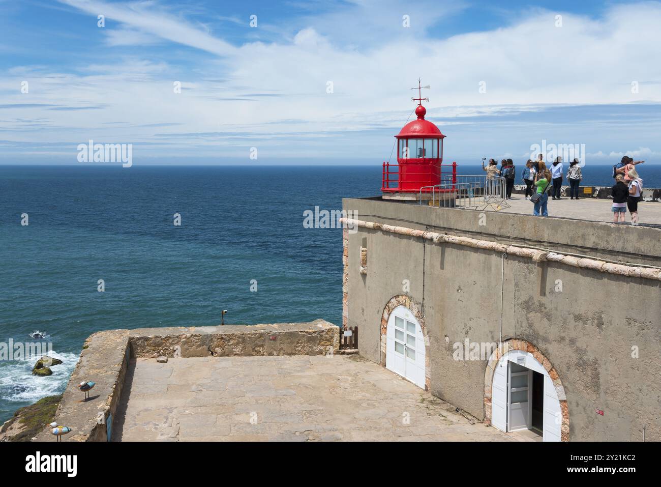 Tourists look at a red lighthouse on a coastal platform with blue sea ...