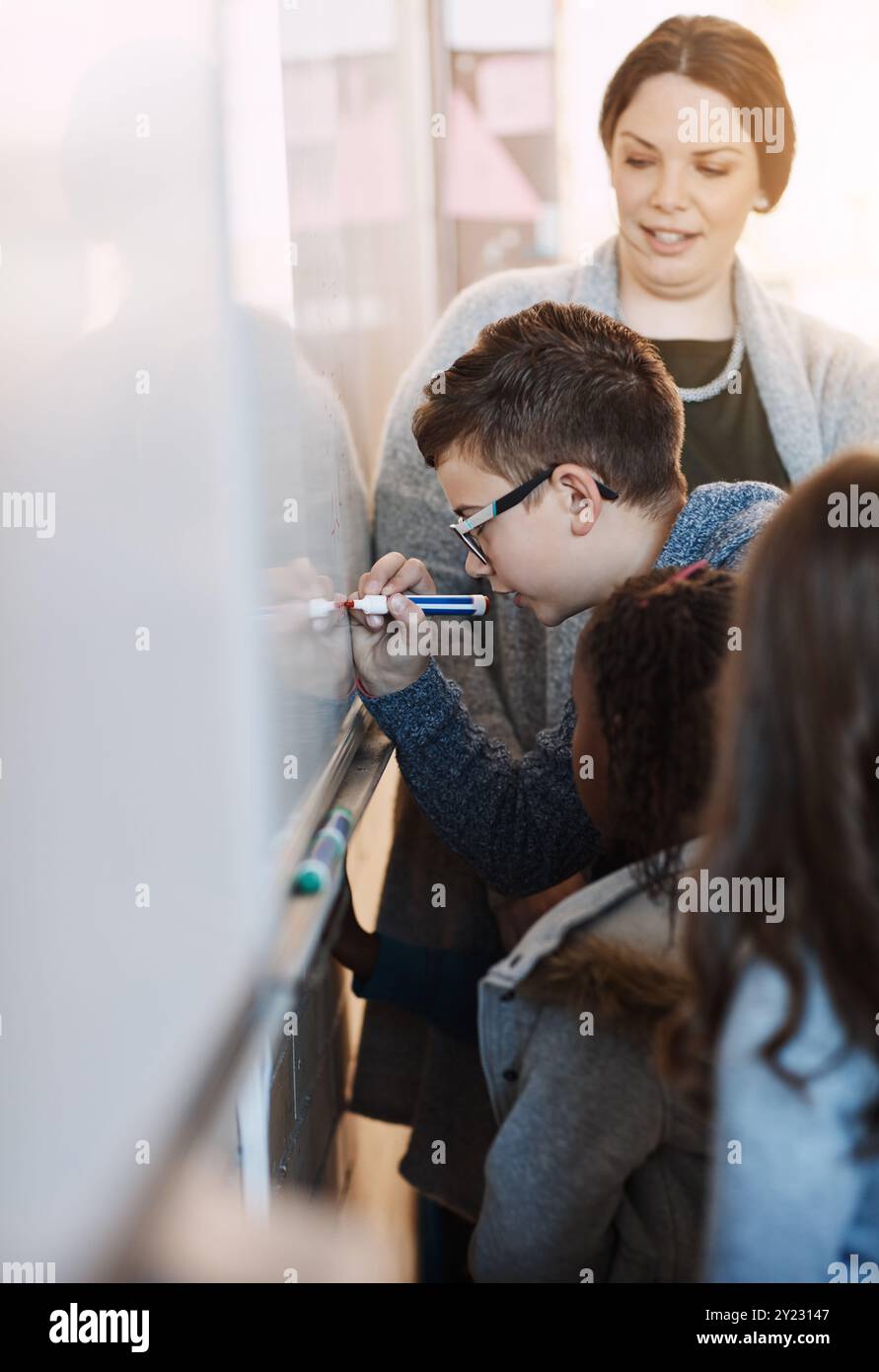 Boy, teacher woman and writing on whiteboard in classroom with children ...