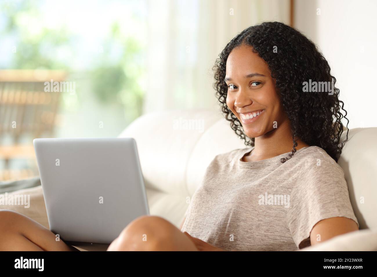 Happy black woman using laptop looks at camera sitting on a couch at ...