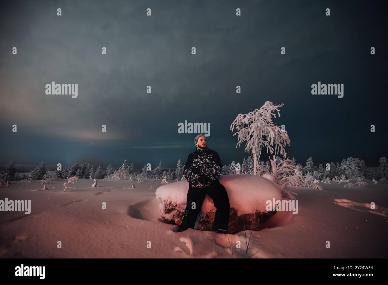 Man sat on snowy rock under faint northern lights in Rovaniemi Lapland ...