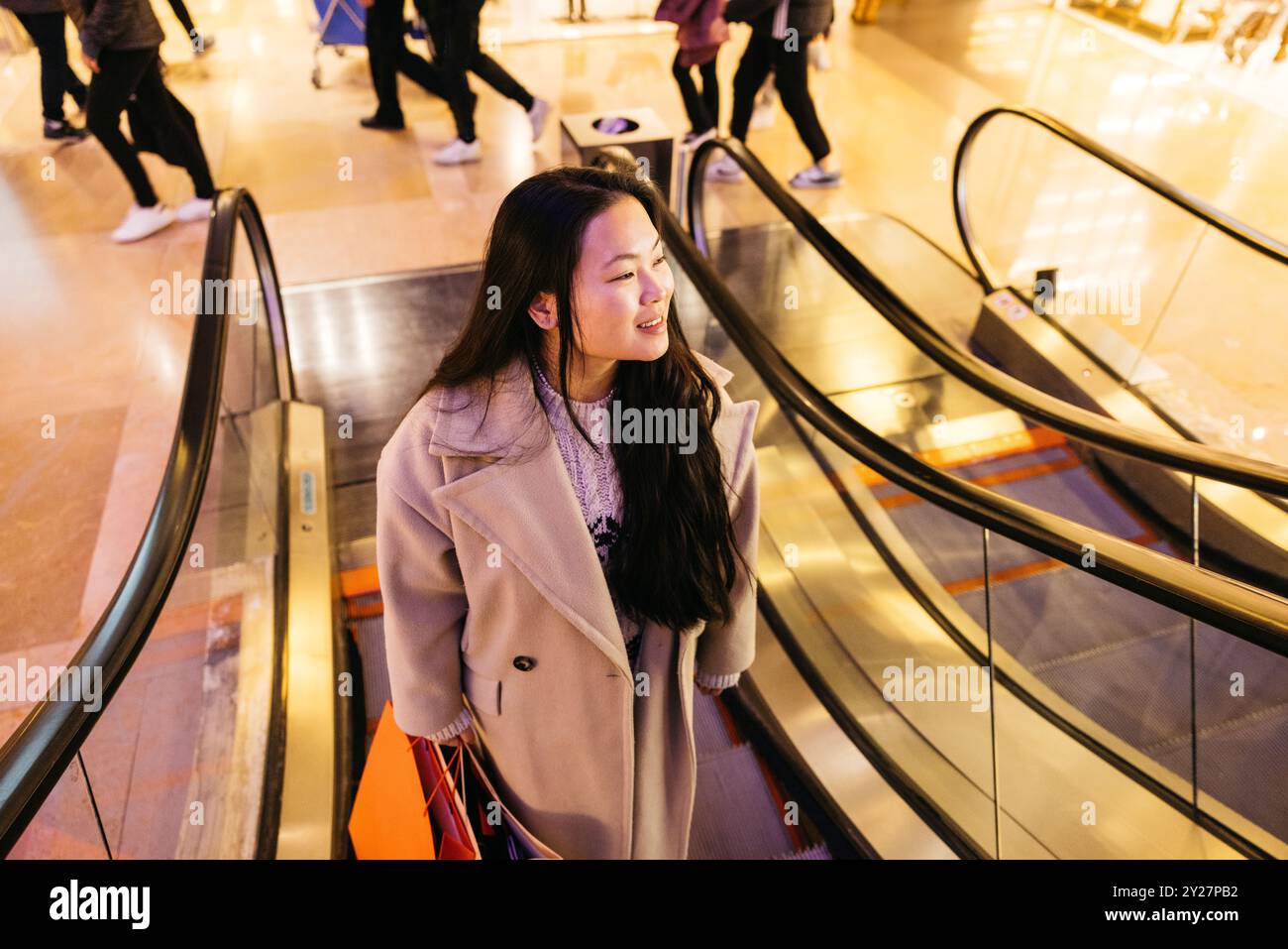 Young Asian woman riding an escalator in a shopping mall, carrying shopping bags, and looking ...