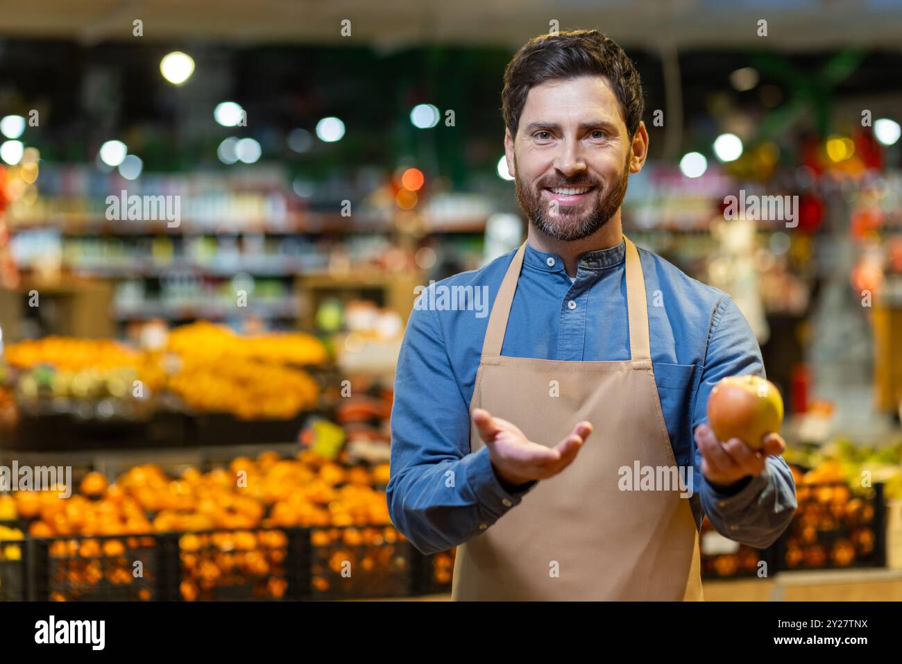 Happy grocery store employee wearing apron holding apple at produce ...
