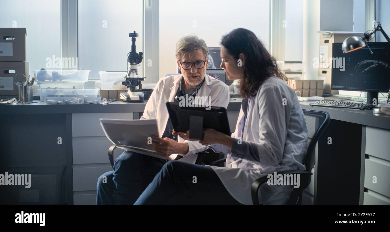 Two diverse scientists in lab coats discuss scientific research, look at papers and use tablet ...