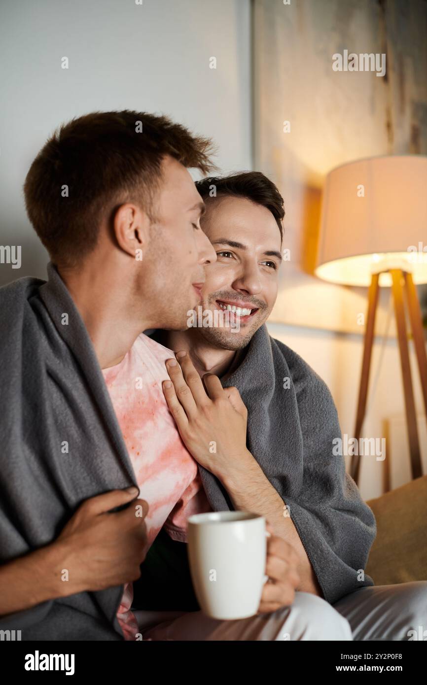 Two men share a joyful moment wrapped in a blanket, sipping drinks ...
