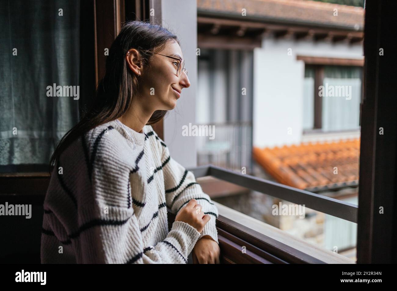 A smiling woman looking away while leaning on a window sill indoors ...