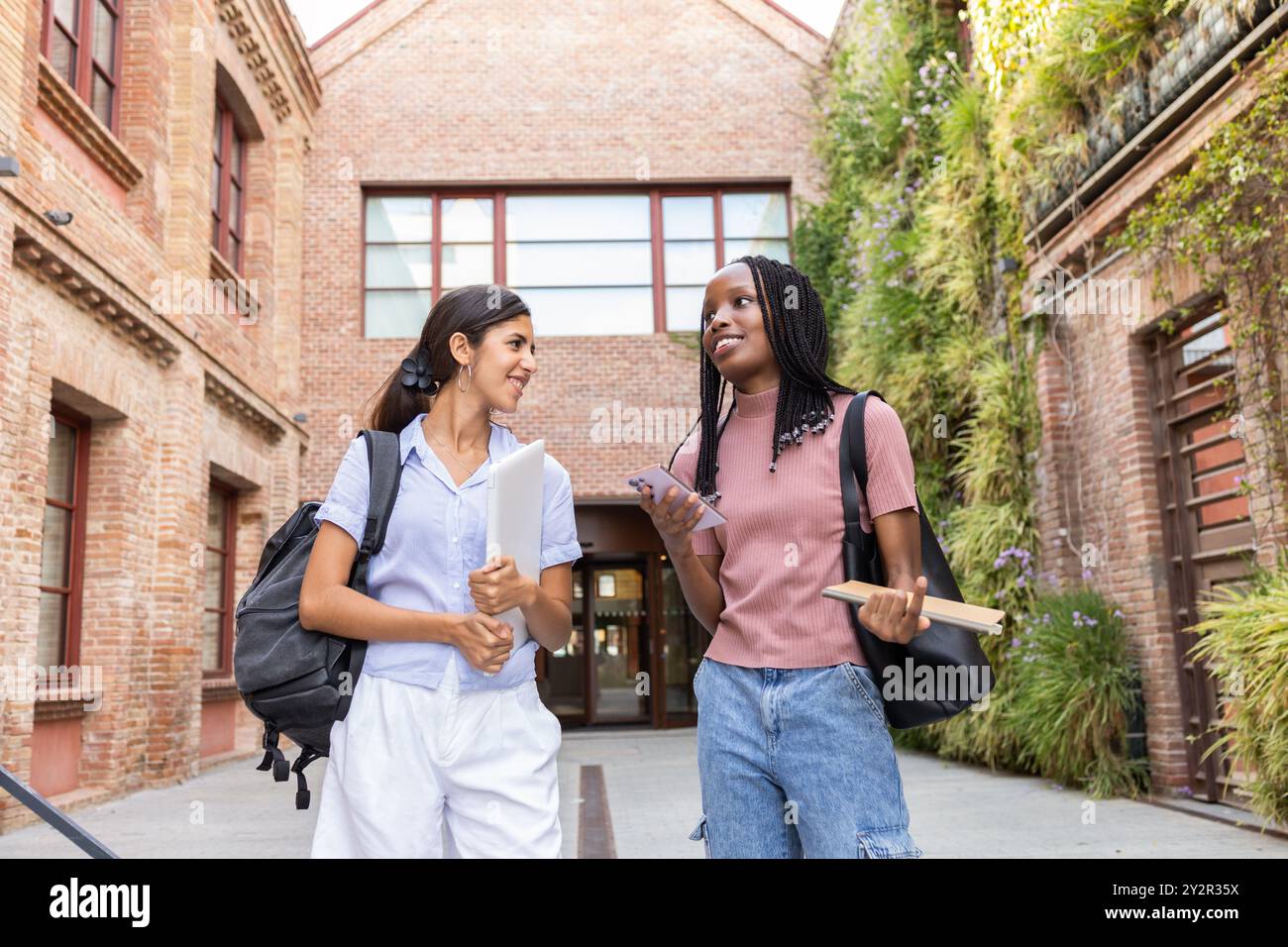Two diverse female students with backpacks walking and chatting happily ...