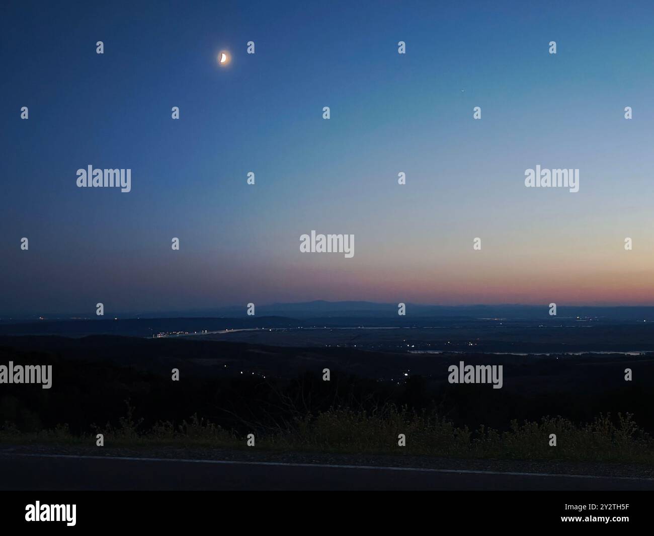 A serene twilight landscape with a crescent moon over a distant horizon ...