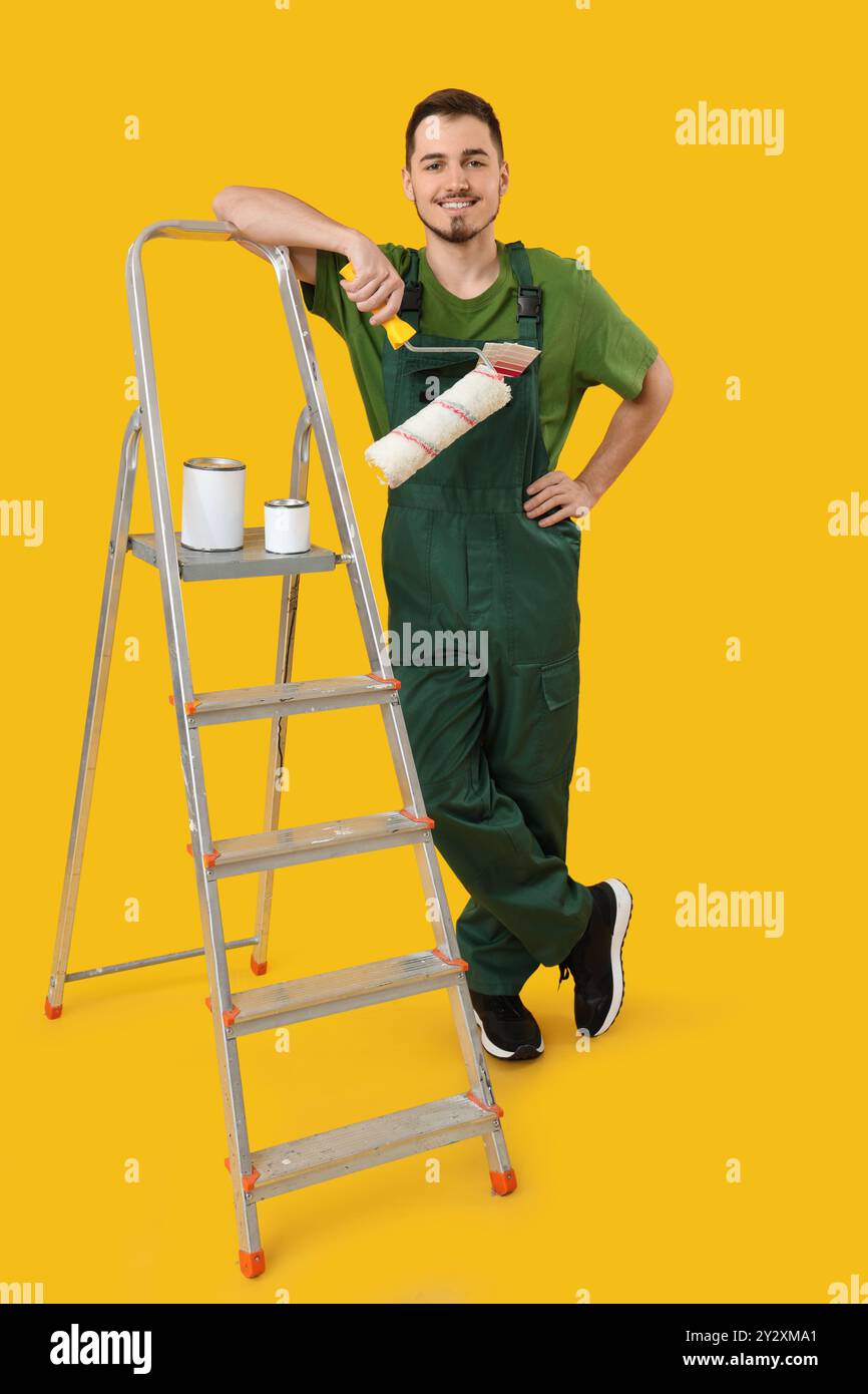Young painter with can of paint, ladder and roller on yellow background ...