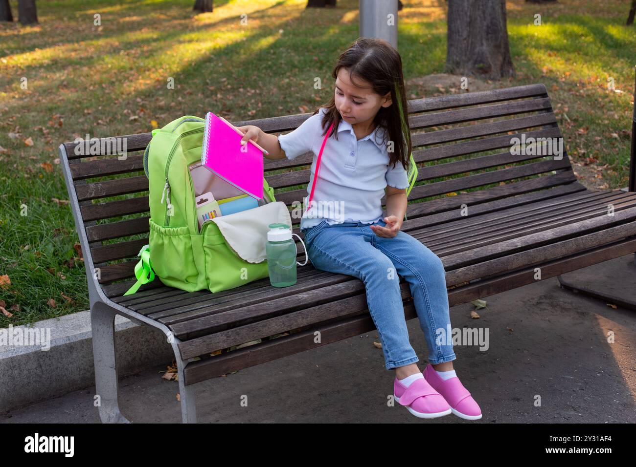 A schoolgirl girl is sitting on a park bench, putting a pink notebook ...